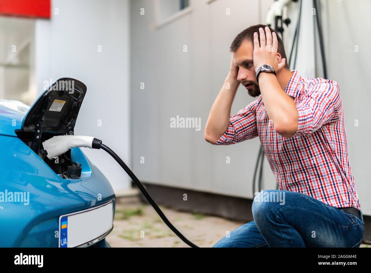 Handsome man plugging cable to electric car with hands on head shocked ...