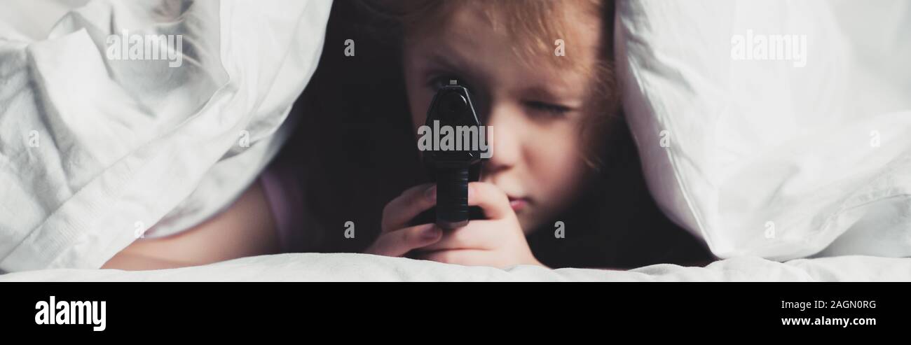 panoramic shot of scared child holding gun while hiding under blanket ...