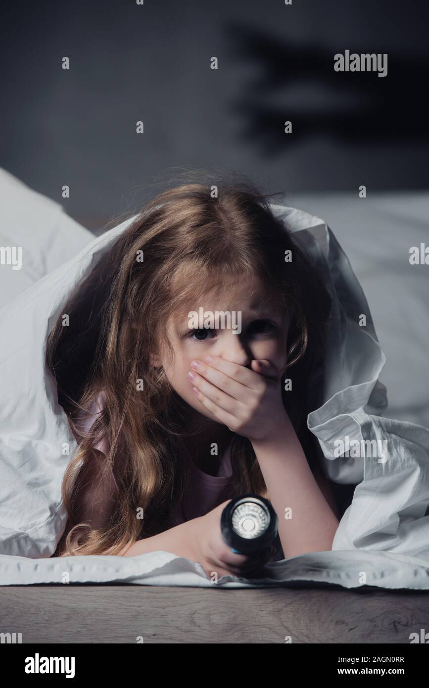 scared child holding flashlight while hiding under blanket on black ...