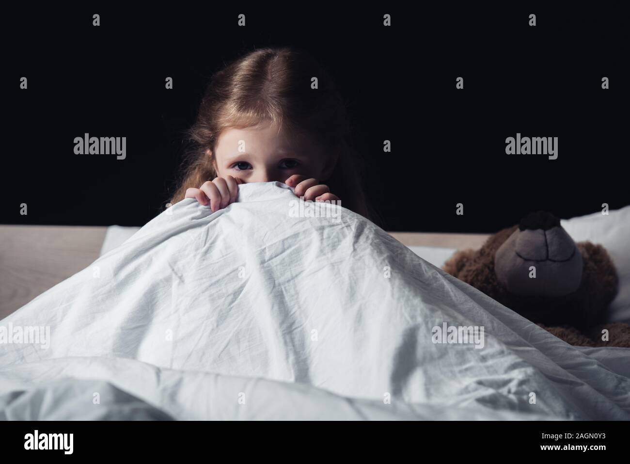 scared child hiding under blanket while sitting on bedding near teddy ...