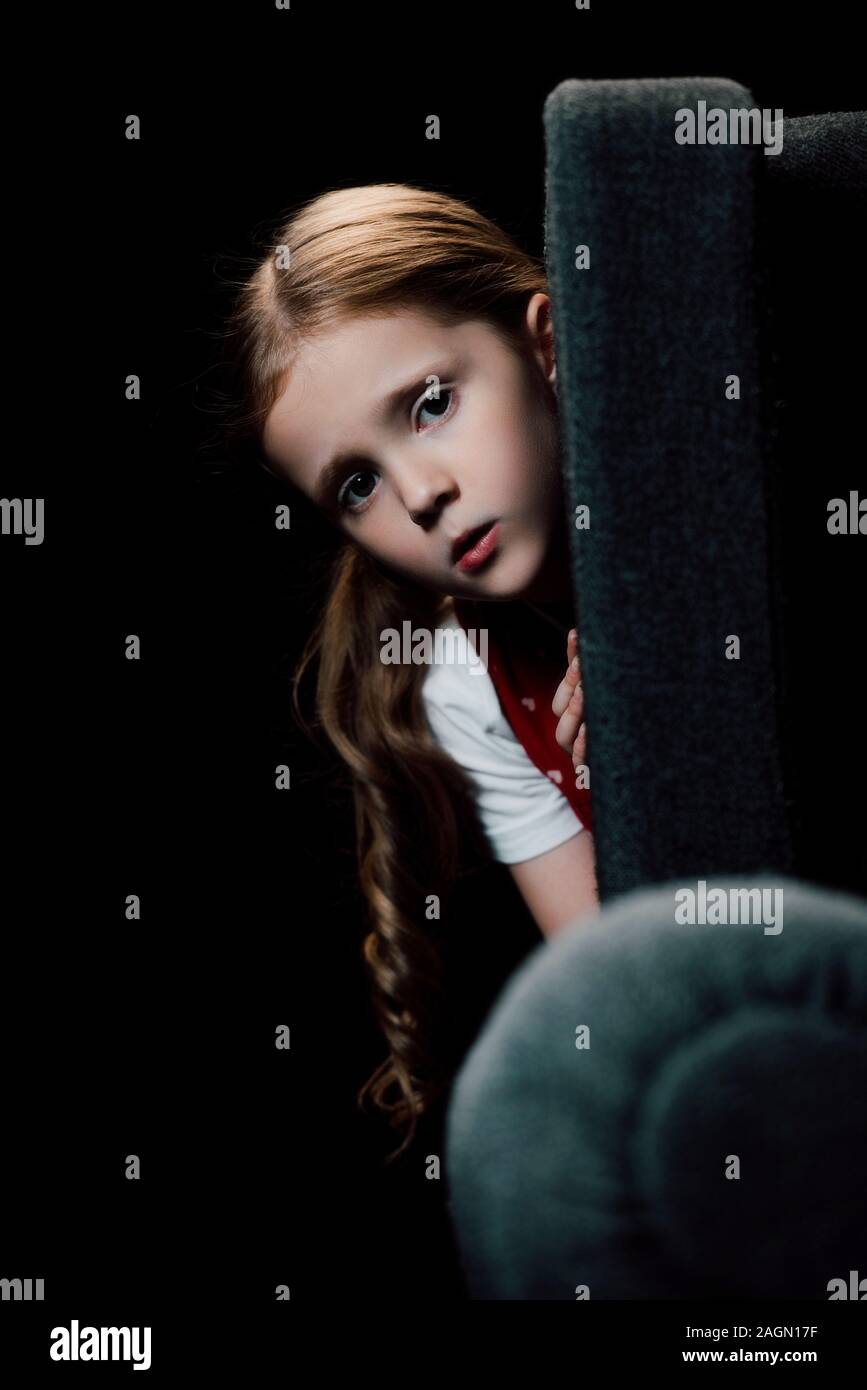 anxious child looking at camera while hiding behind armchair isolated ...