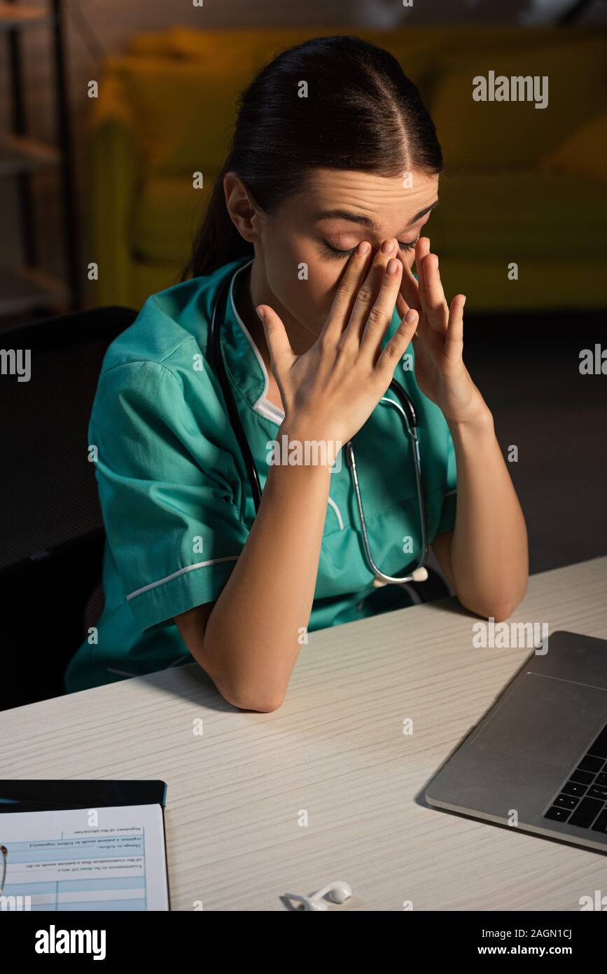 attractive and tired nurse in uniform sitting at table during night ...