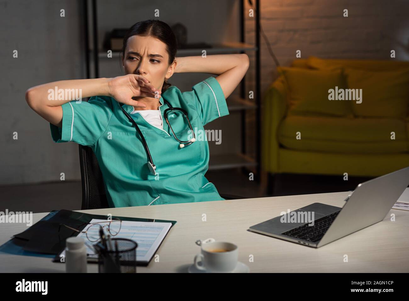 attractive nurse in uniform sitting at table and yawning during night ...