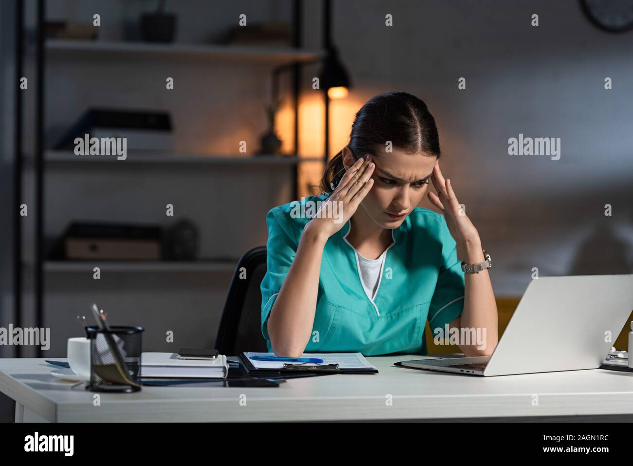 attractive and tired nurse in uniform sitting at table during night ...