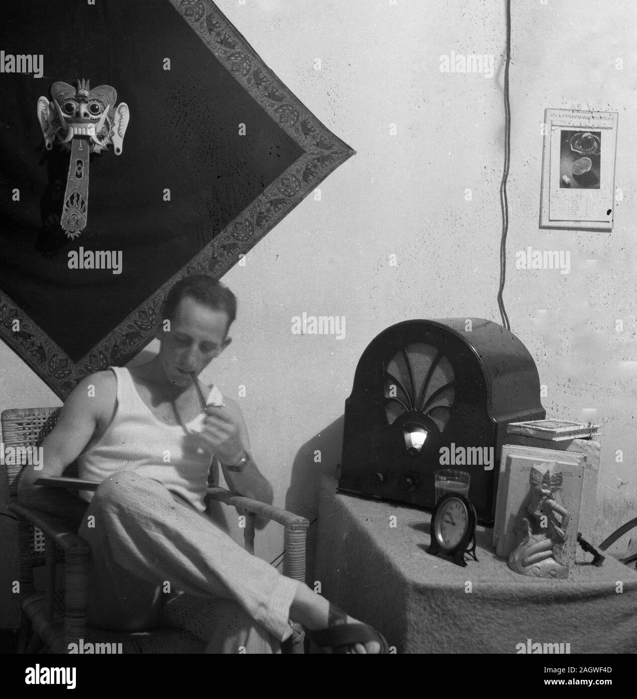Man in a room smokes a pipe while reading. On the right a table with ...
