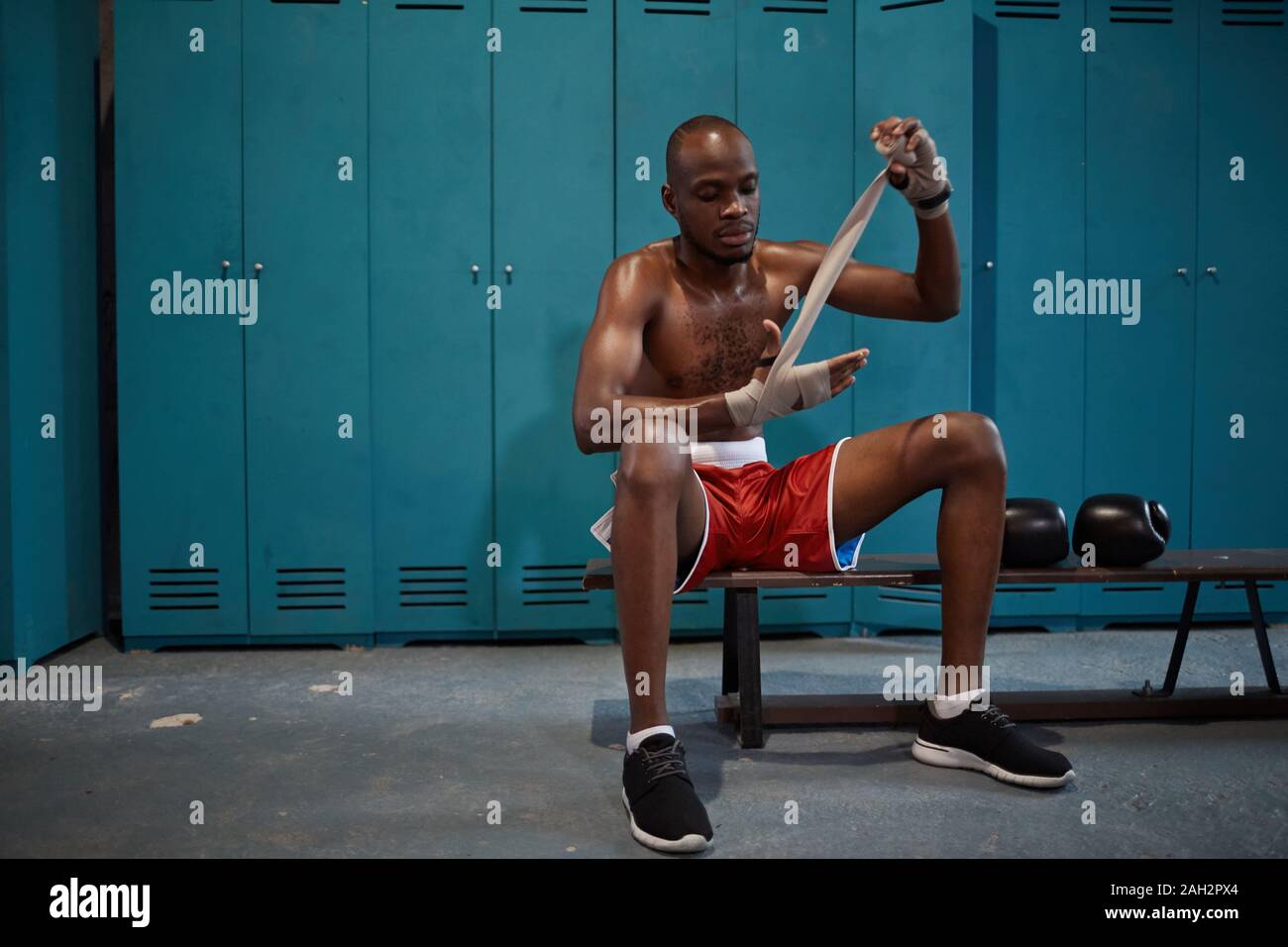 African young boxer sitting on bench and wrapping a bandage around his ...