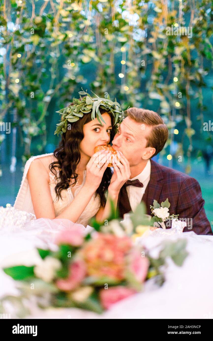 Close up of funny happy wedding couple eating burger, sitting in ...