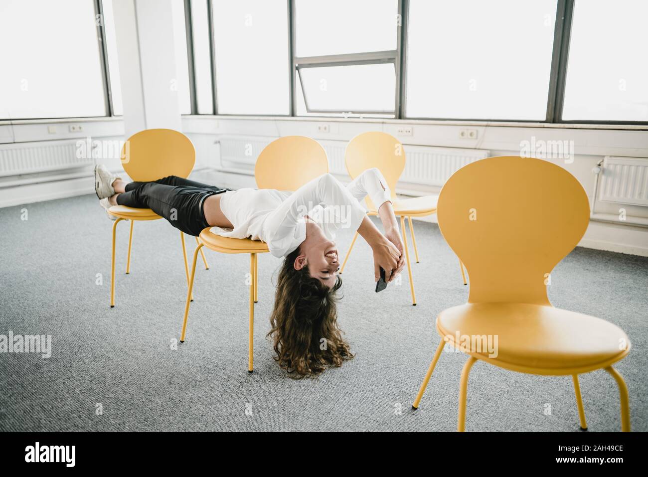 Smiling businesswoman lying across chairs in an empty office using ...