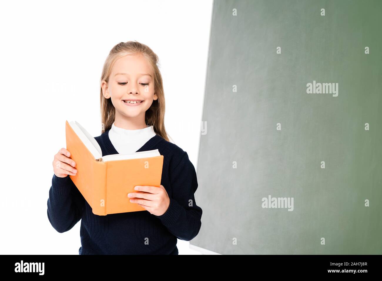cheerful schoolgirl reading book while standing near chalkboard ...