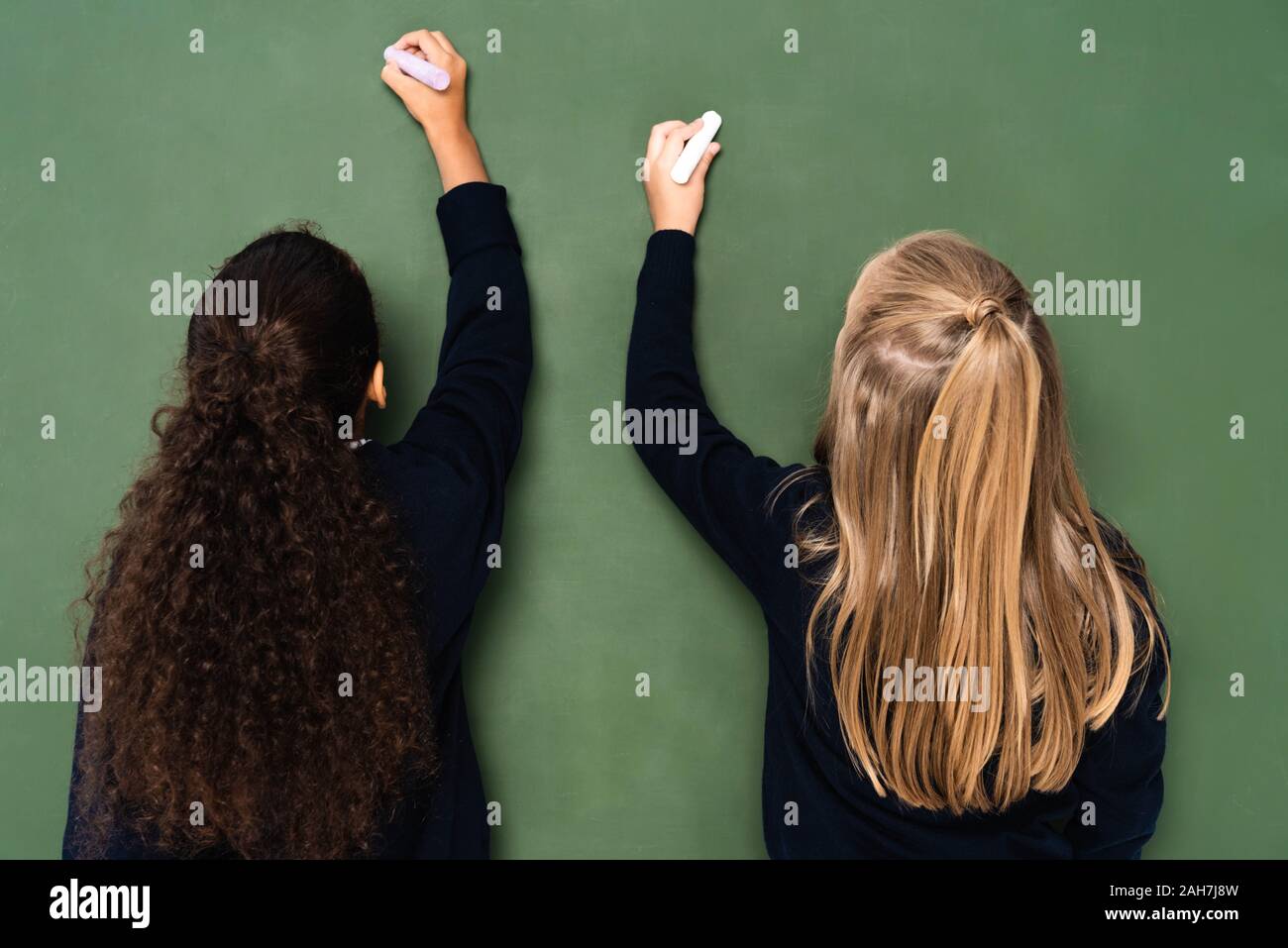 back view of two multicultural schoolgirls writing on chalkboard with ...