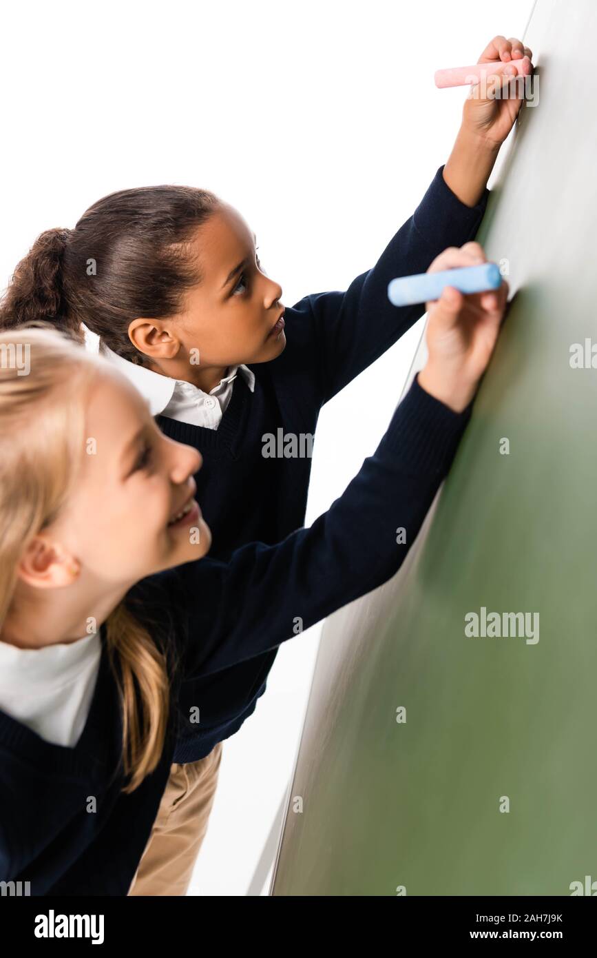 two adorable multicultural schoolgirls writing on green chalkboard ...