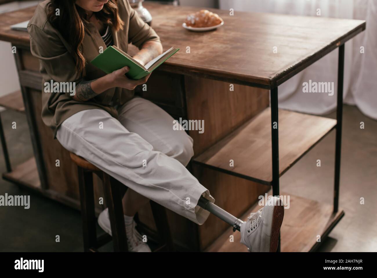 Cropped view of girl with leg prosthesis reading book beside table at ...