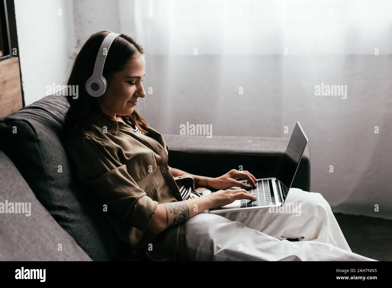 Side view of smiling freelancer in headphones working at laptop on sofa ...