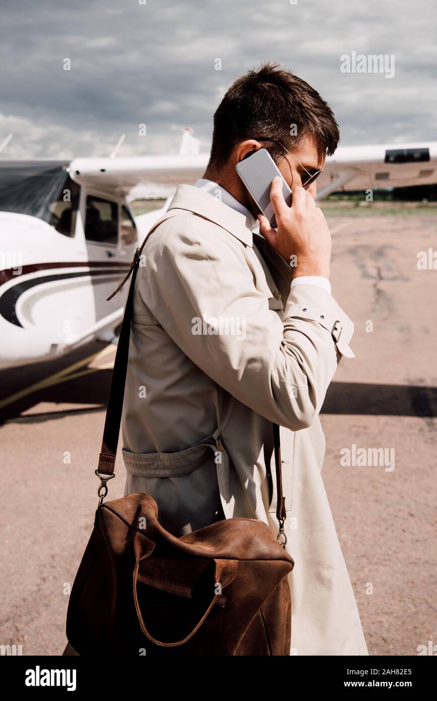 side view of man in coat with bag talking on smartphone near plane ...