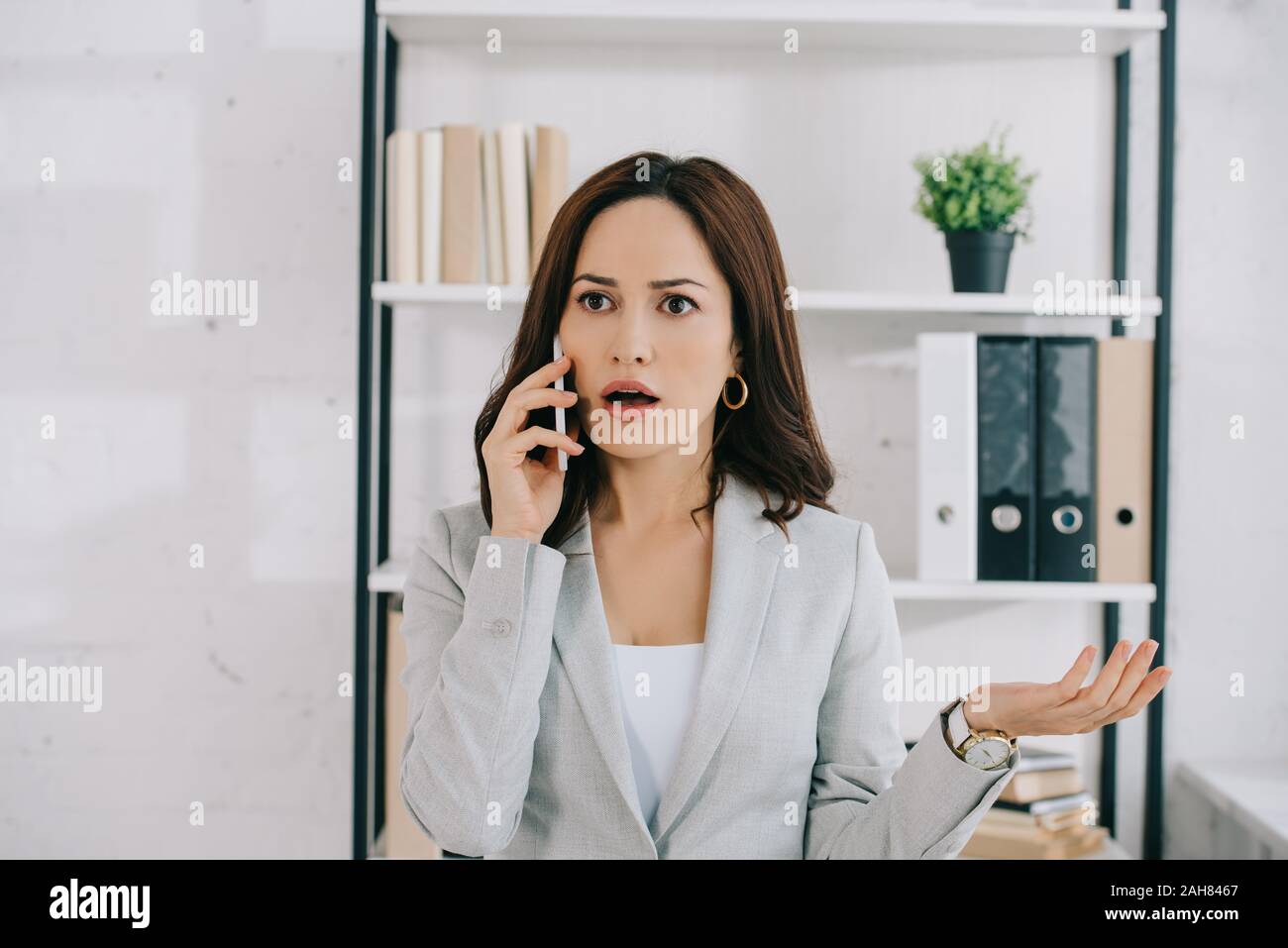 shocked secretary showing shrug gesture while talking on smartphone in ...