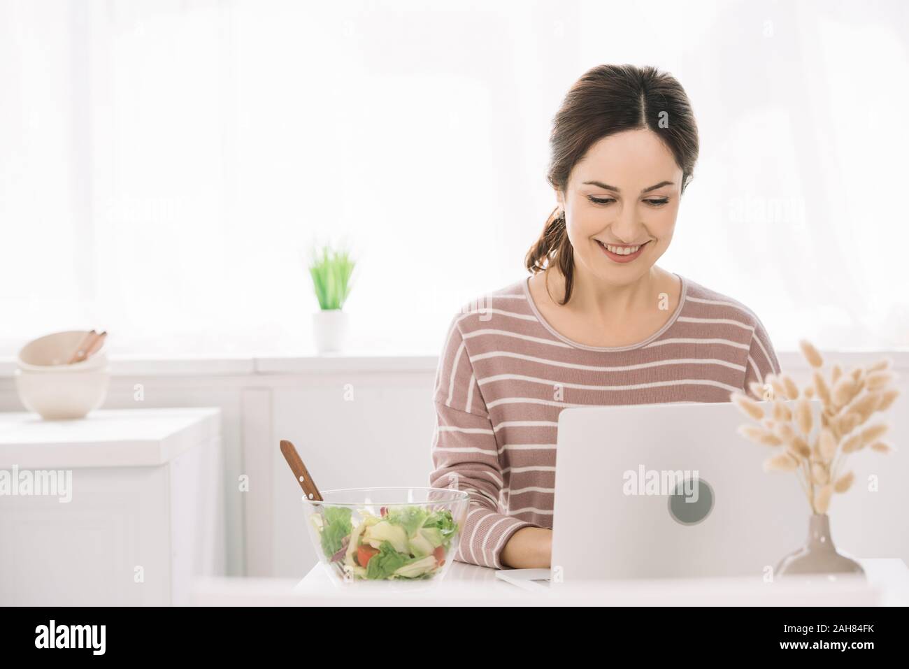 young, happy woman using laptop while sitting at kitchen table near ...