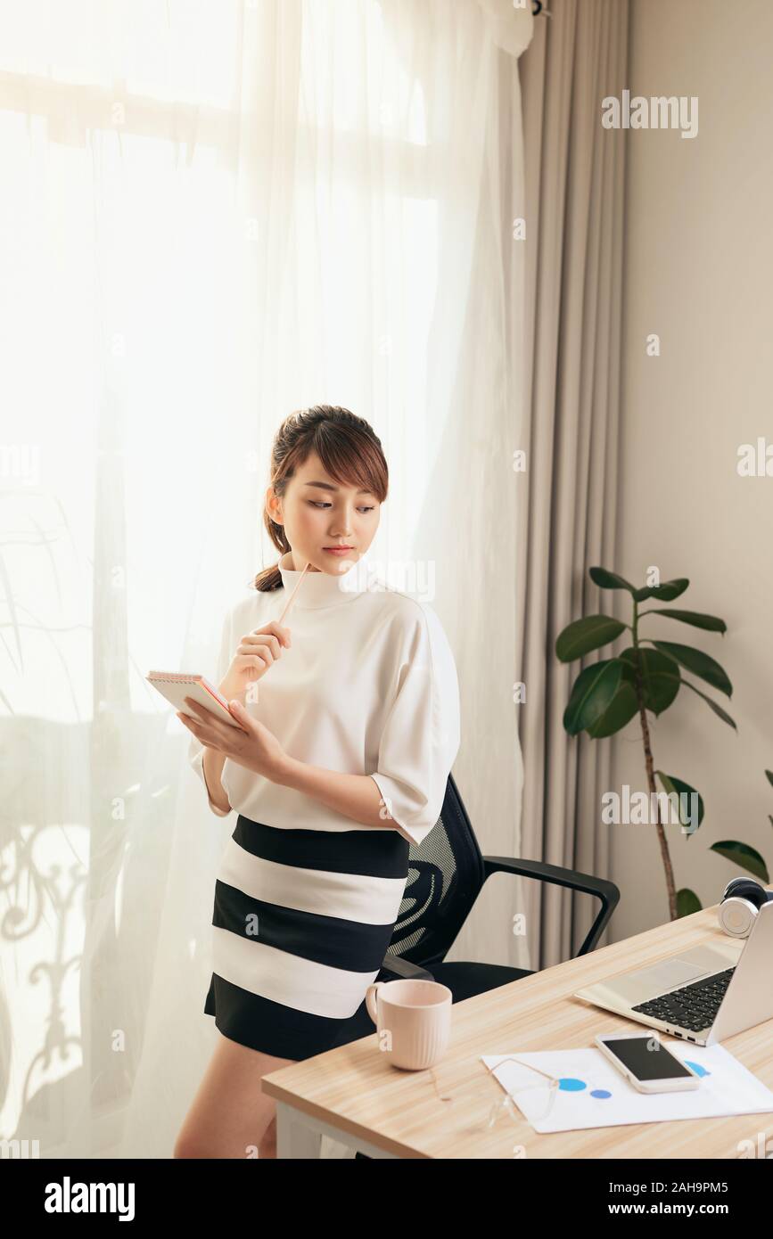 Young Asian woman standing to write in notebook while looking at laptop ...
