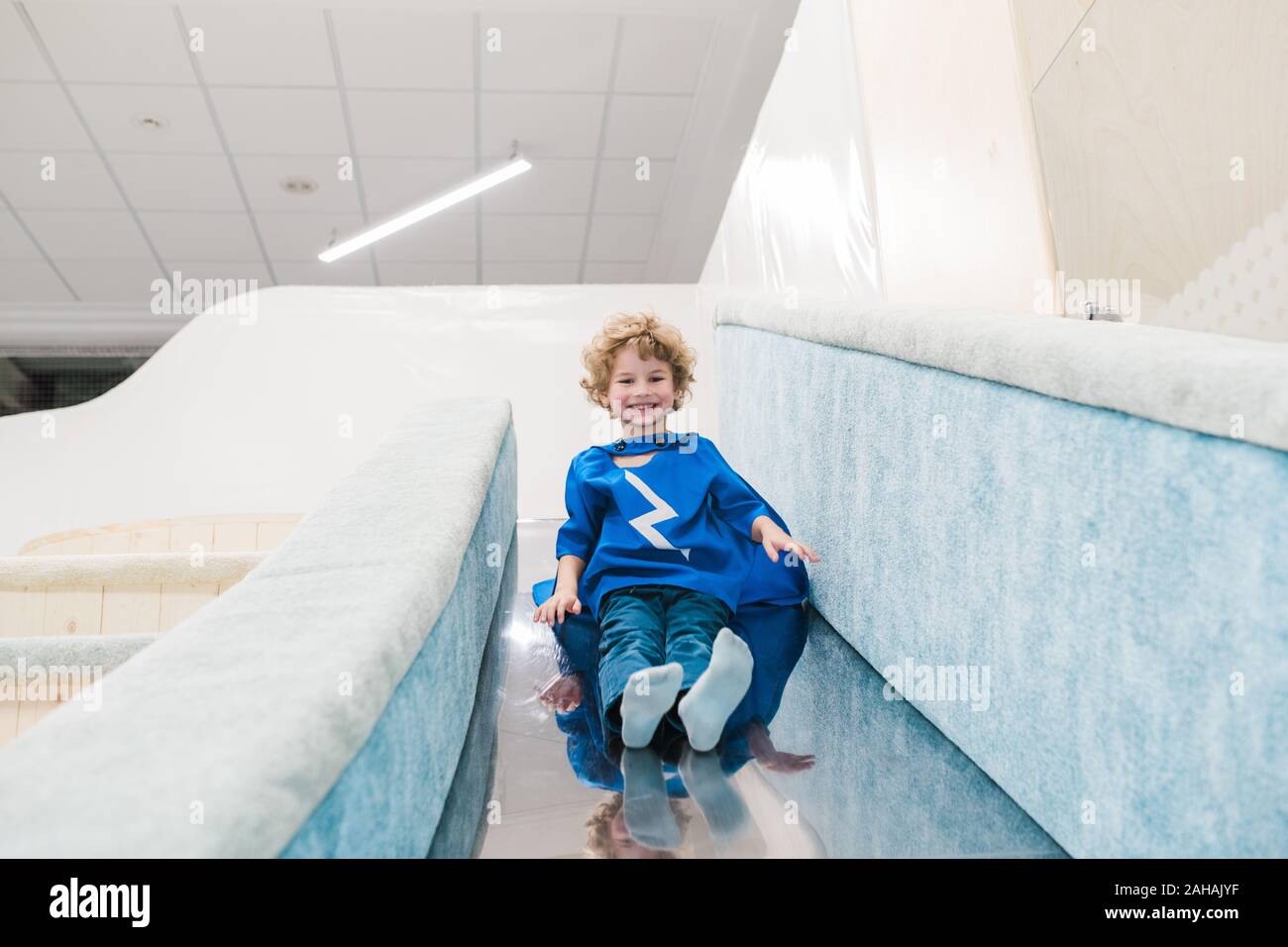 Cute cheerful little boy in blue costume of superman moving down chute ...