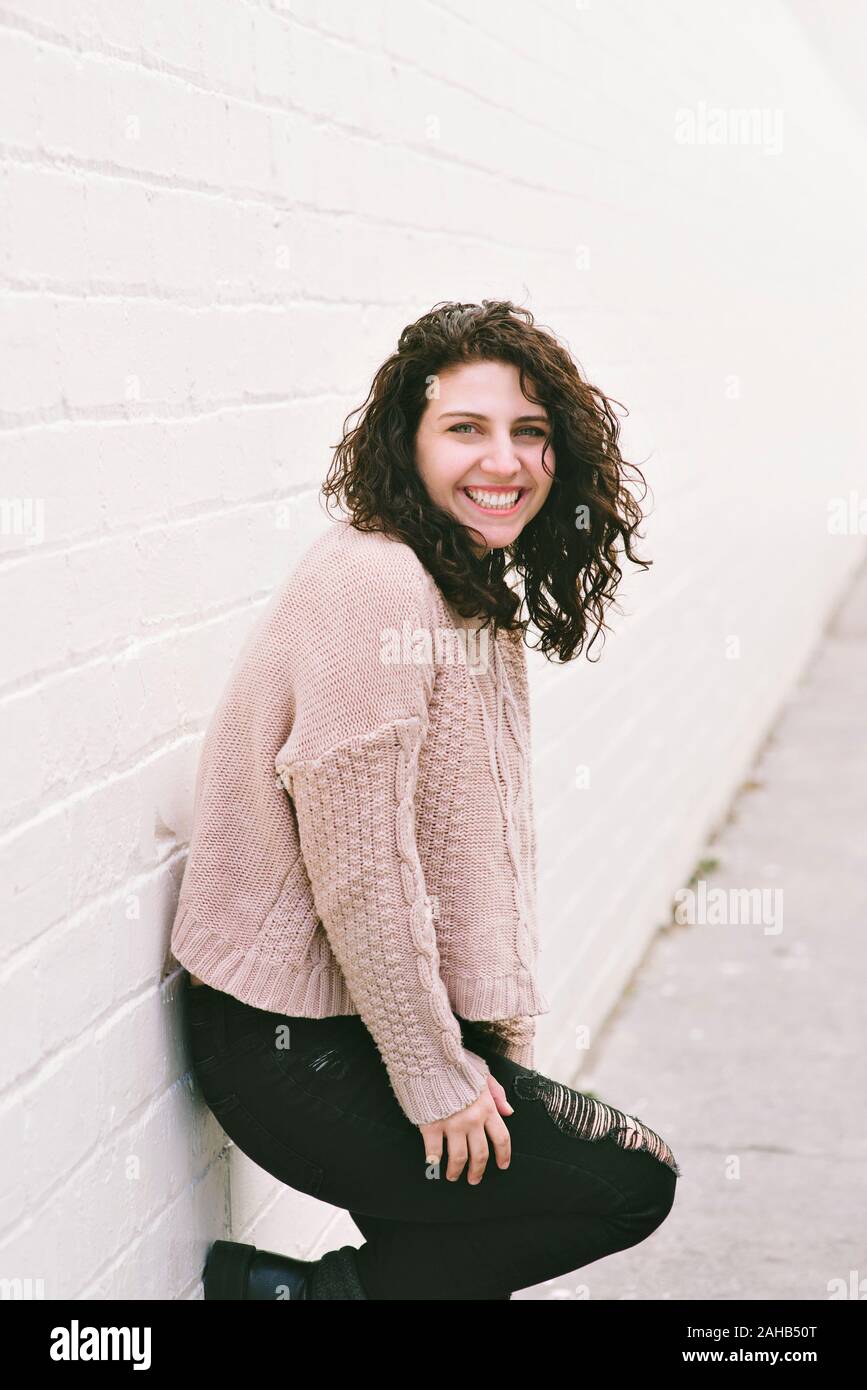 Young female millennial leans against a wall with a big smile and ...