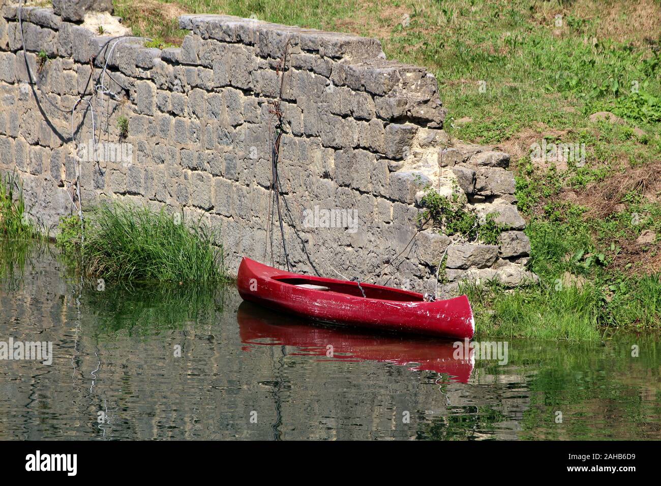 Dilapidated dark red plastic canoe boat tied to old broken stone wall ...