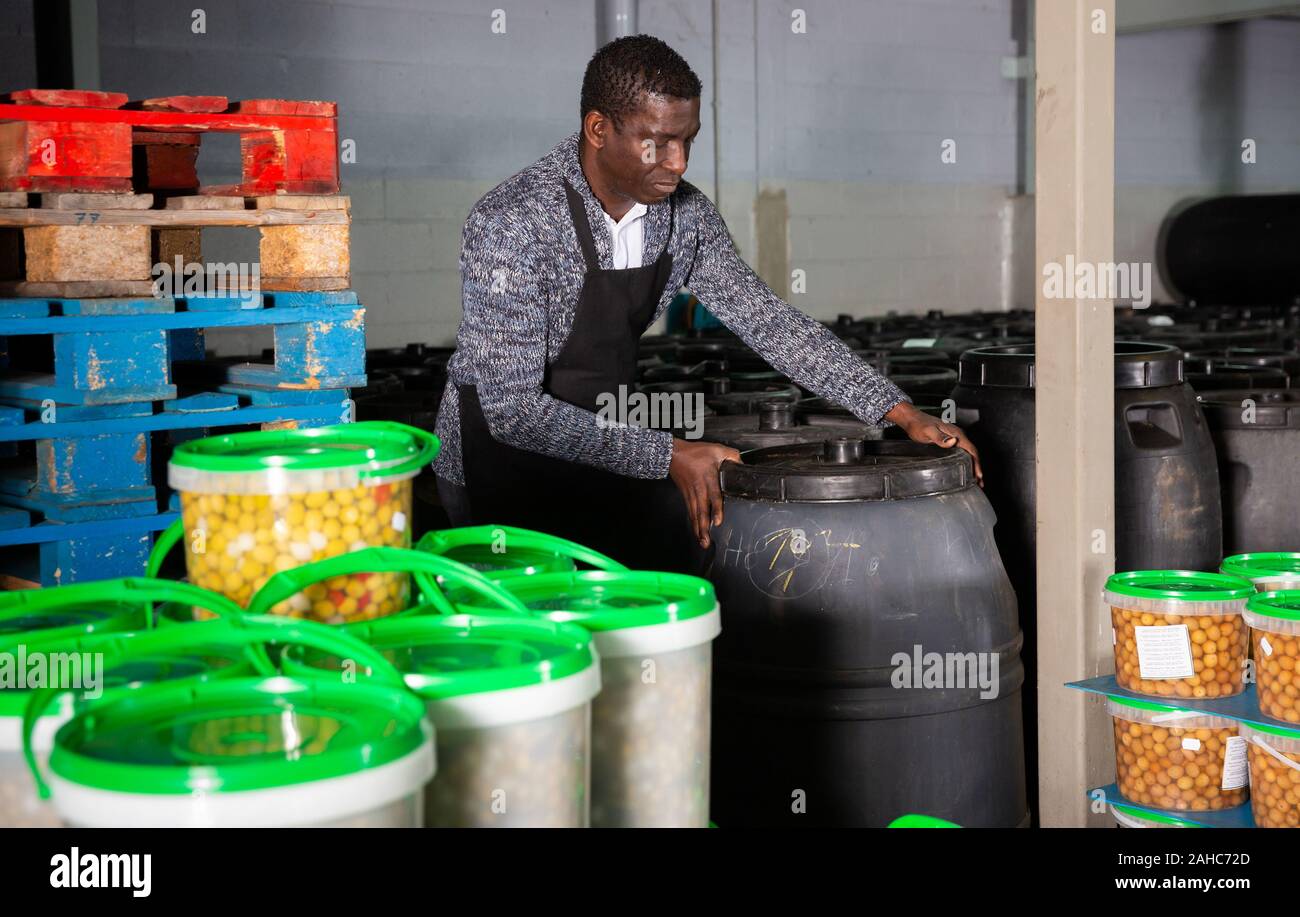 African man worker of pickled olives factory stacking plastic barrels ...