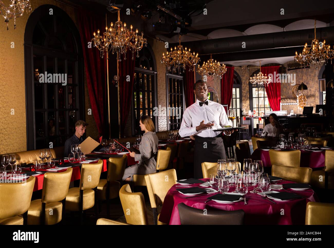 Polite waiter holding tray in restaurant with customers behind him ...