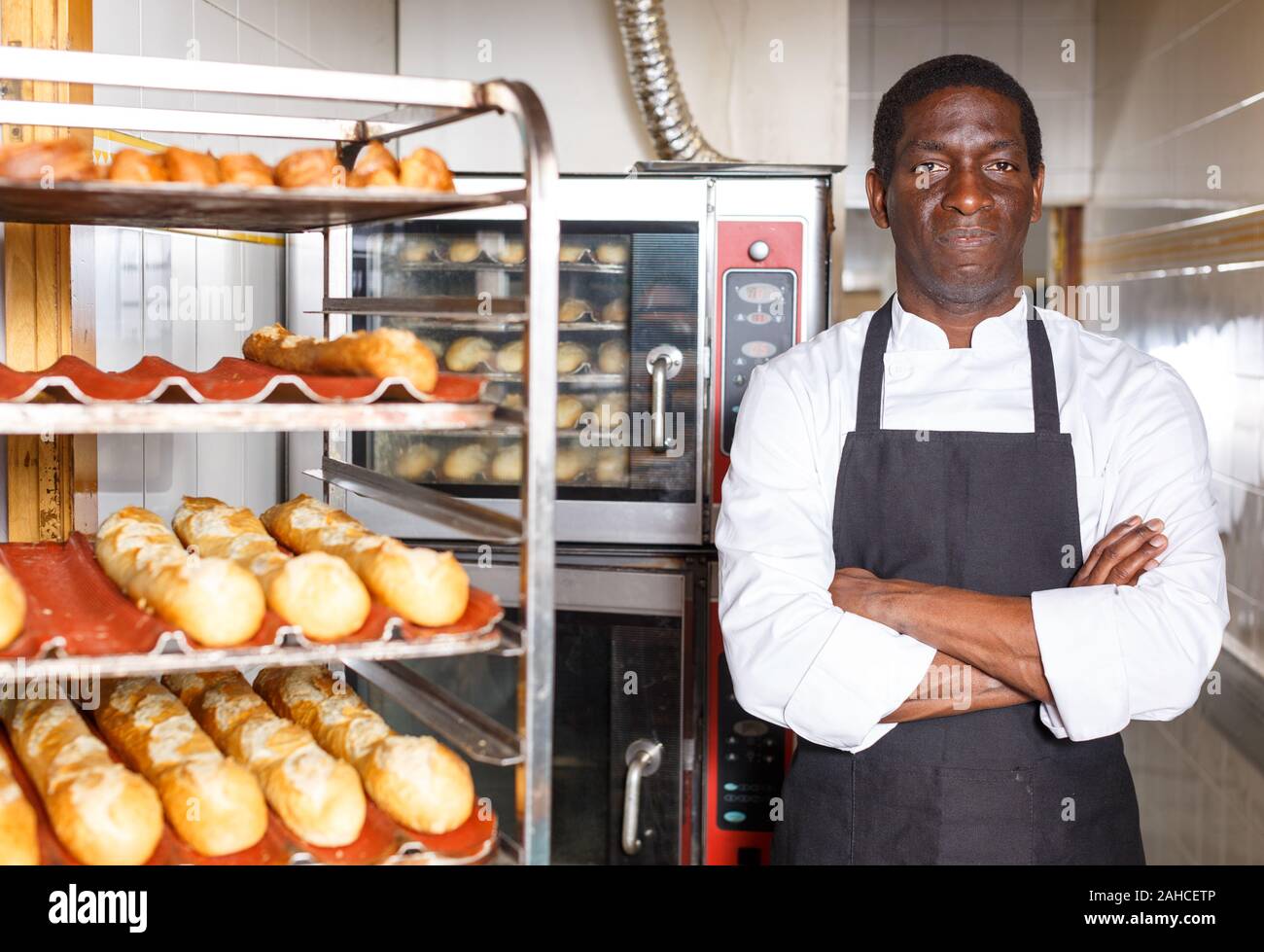 Confident baker in black apron standing with arms crossed near professional bread oven in small ...