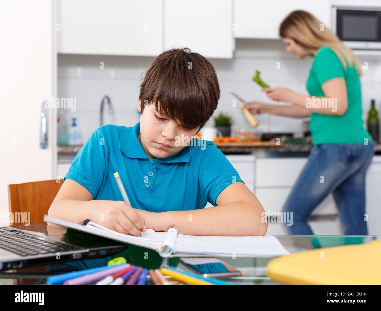 Positive schoolboy doing homework using laptop at kitchen interior ...