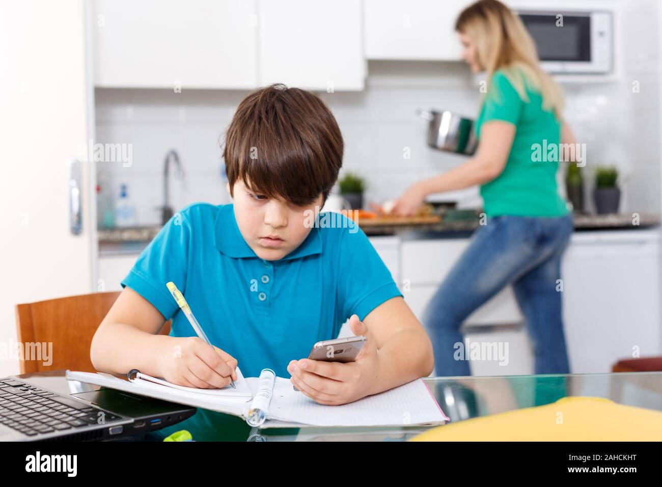 Positive schoolboy doing homework using laptop at kitchen interior ...