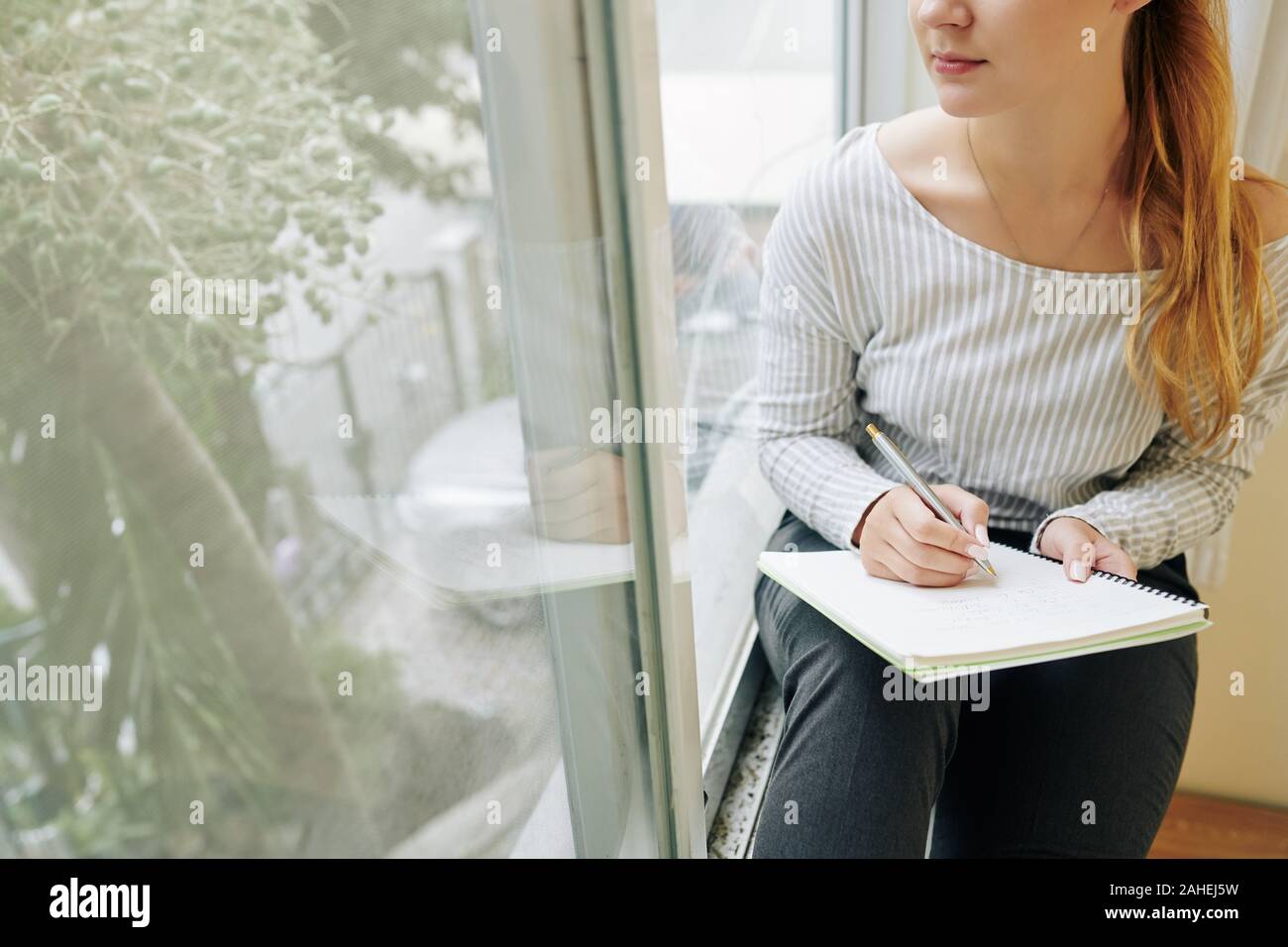Cropped image of pensive young woman sitting on window sill and writing ...