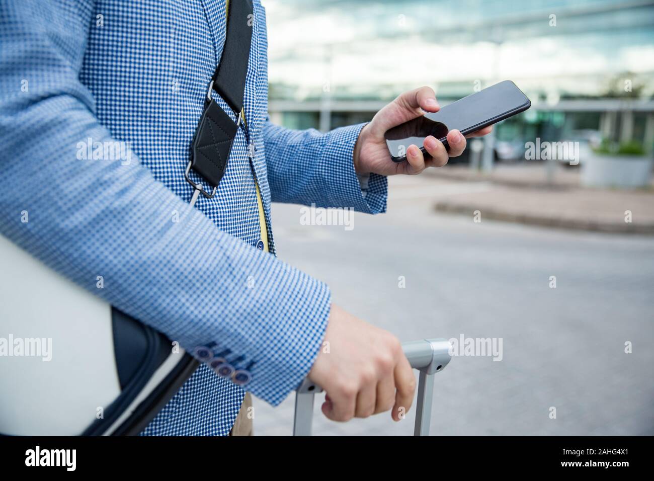 Stylish man standing at airport with smartphone and suitcase, browsing ...