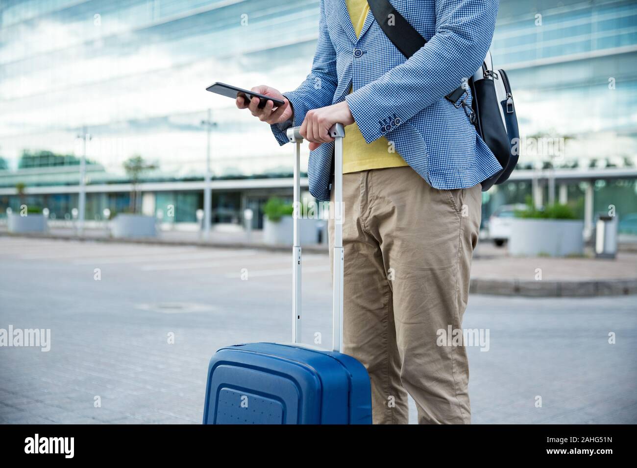 Stylish man standing at airport with smartphone and suitcase, browsing ...