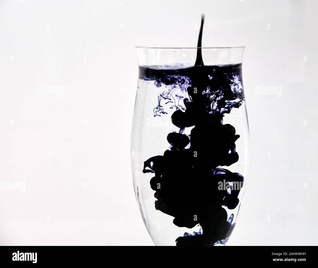 Blue and black ink dropping into a wine glass of water on a white ...