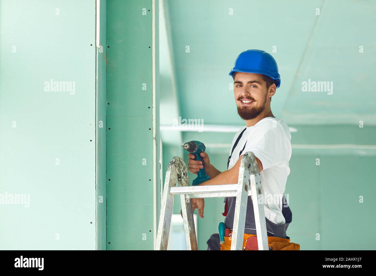 A builder standing on a ladder installs drywall at a construction site ...
