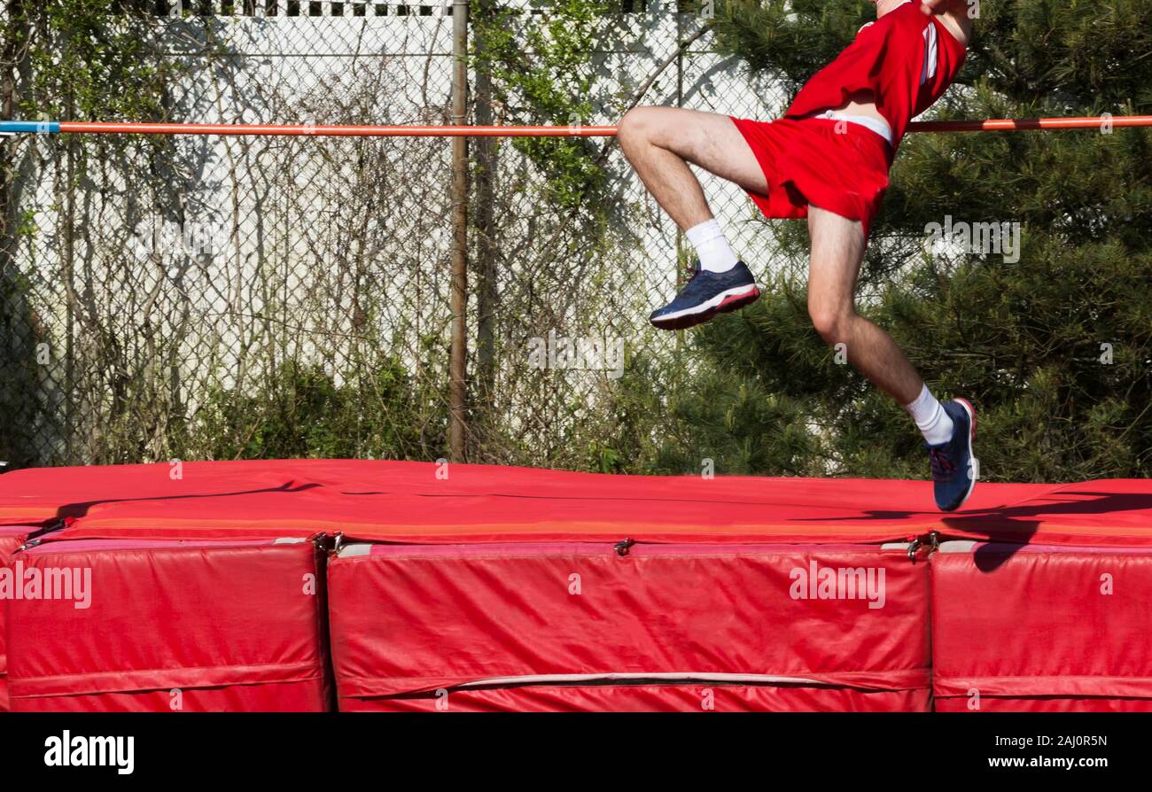 High school boy wearing a red uniform is high jumping on to red mats ...
