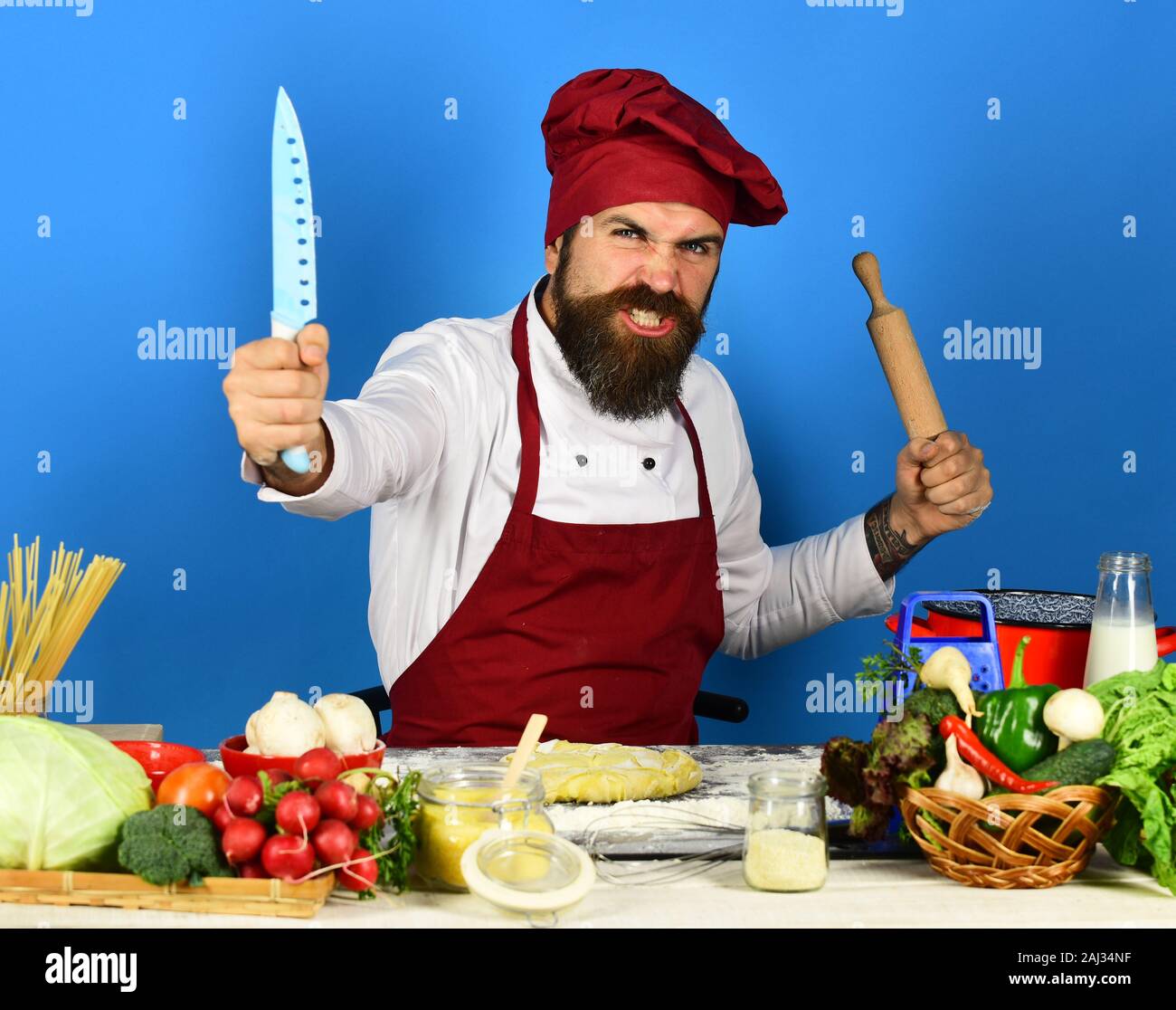 Chef with pasta, vegetables and dough on table. Man with beard sits by ...