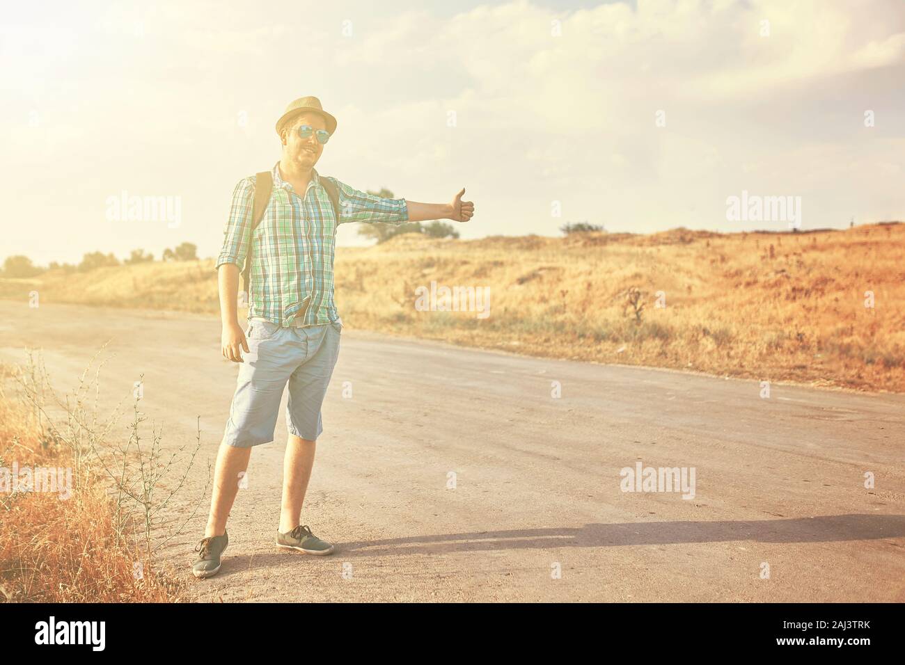 Hitchhiker with backpack in summer hat, light shirt, shorts on the road ...