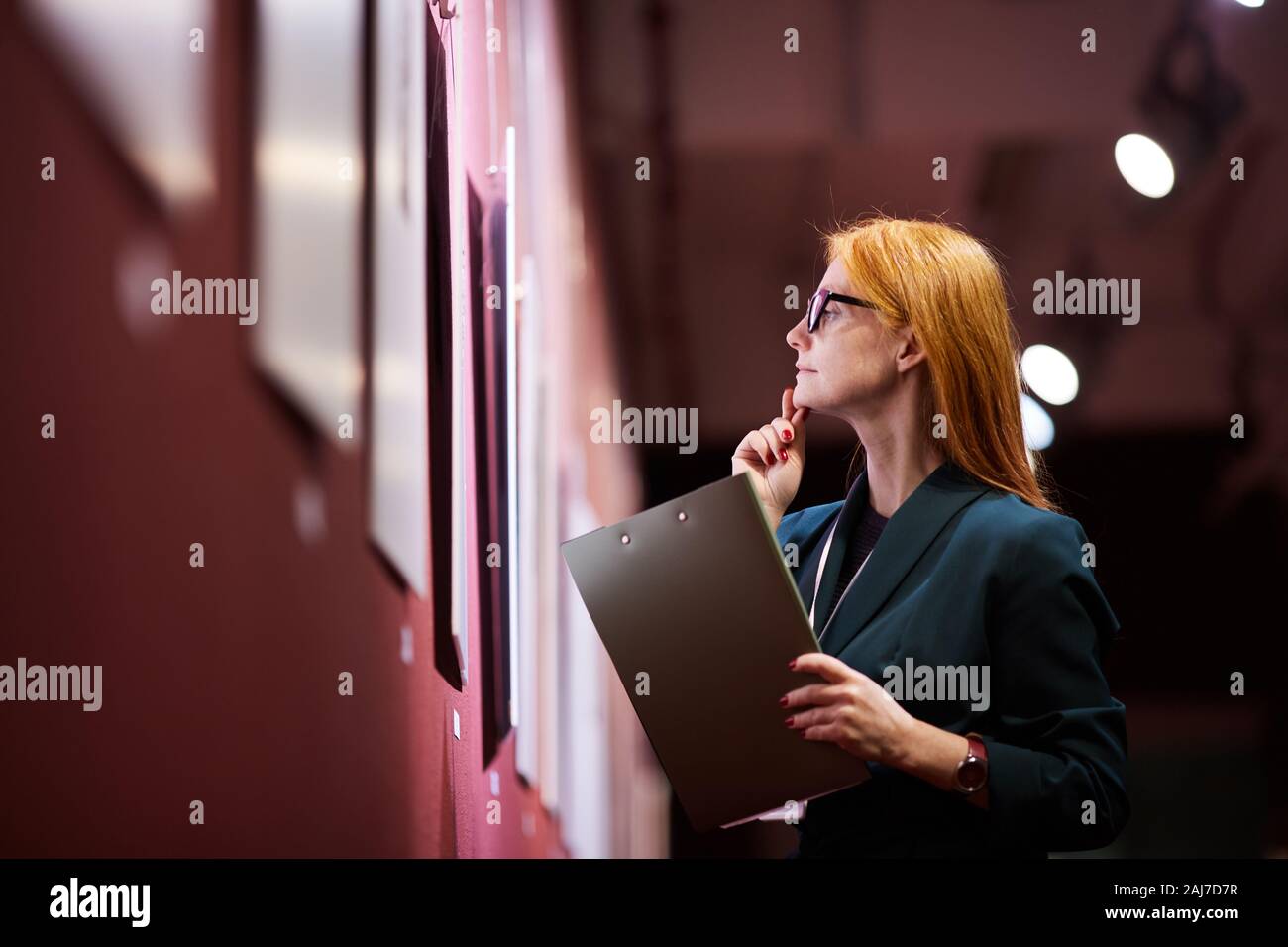 Young serious art manager in eyeglasses holding folder and standing in ...