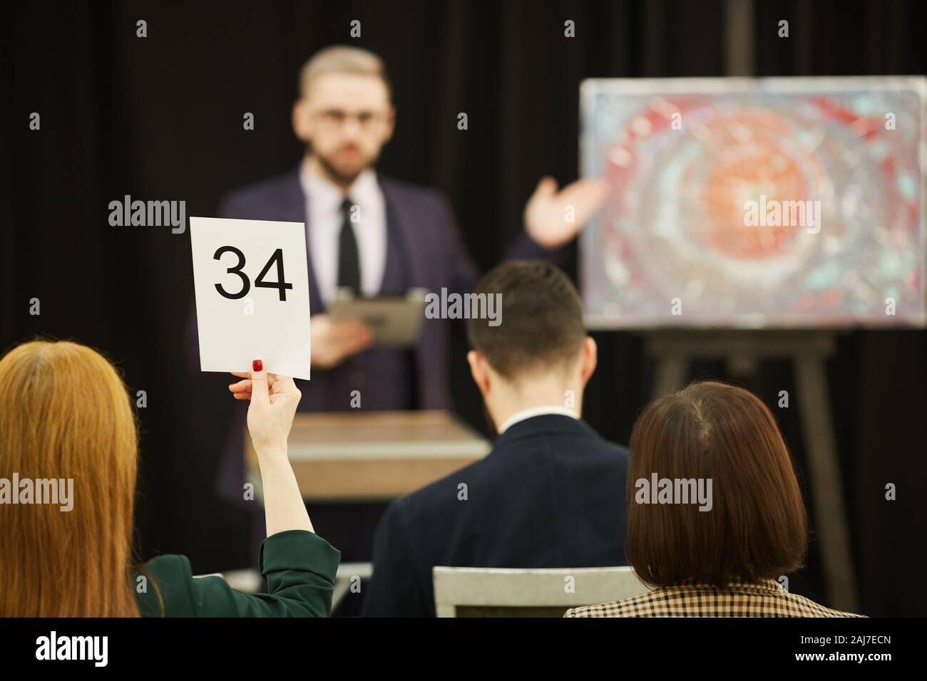 Rear view of group of people sitting at auction and woman raising her ...