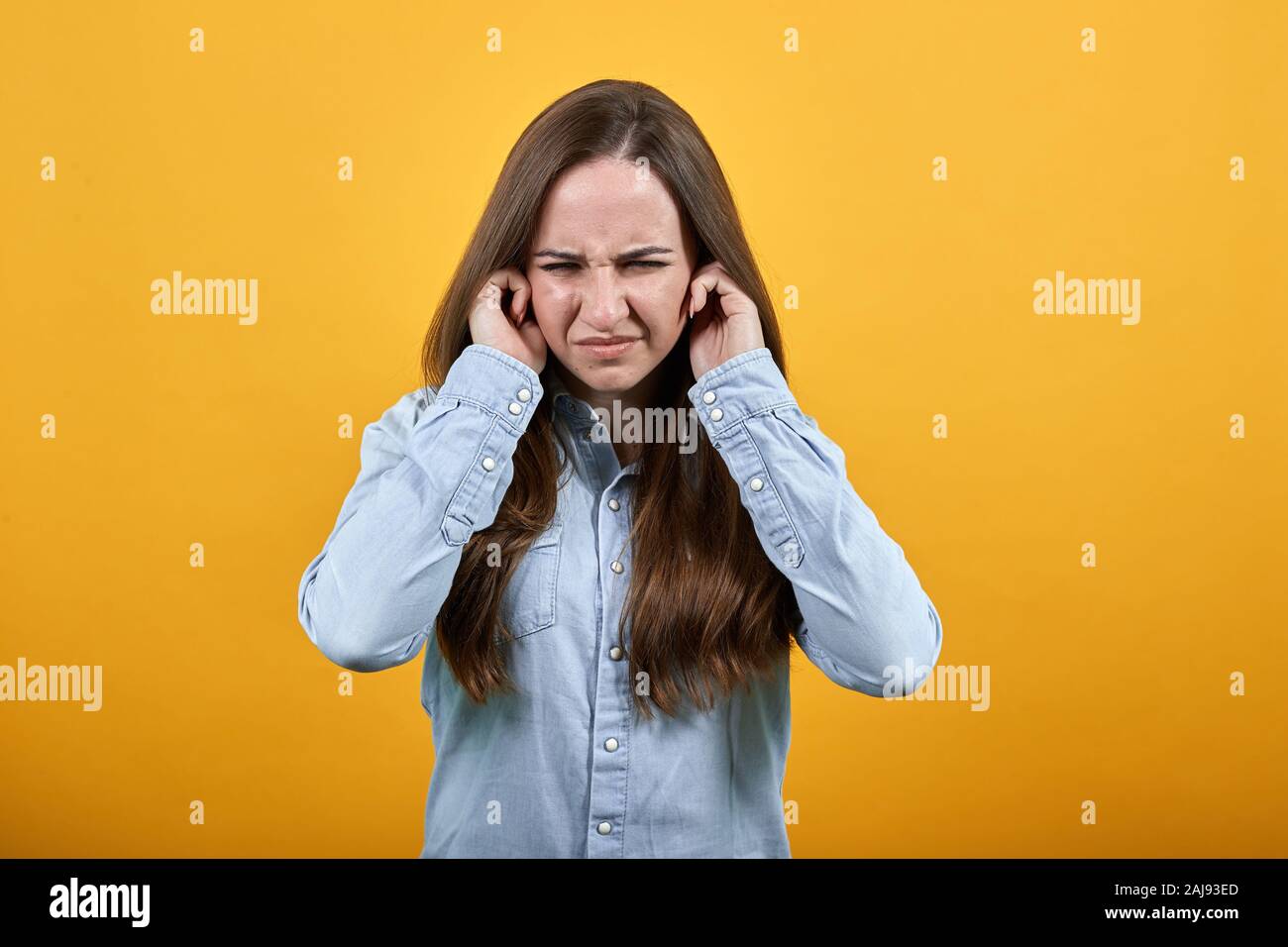 Irritated woman in denim shirt keeping fingers on ears, squeezing face ...