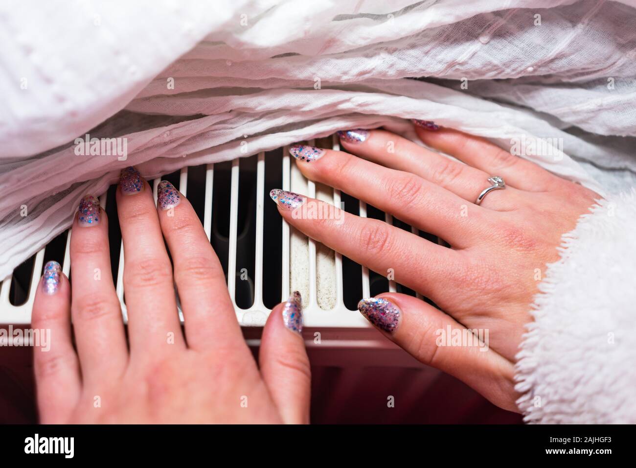 Close-up of a woman warming up her hands on a white radiator at home ...