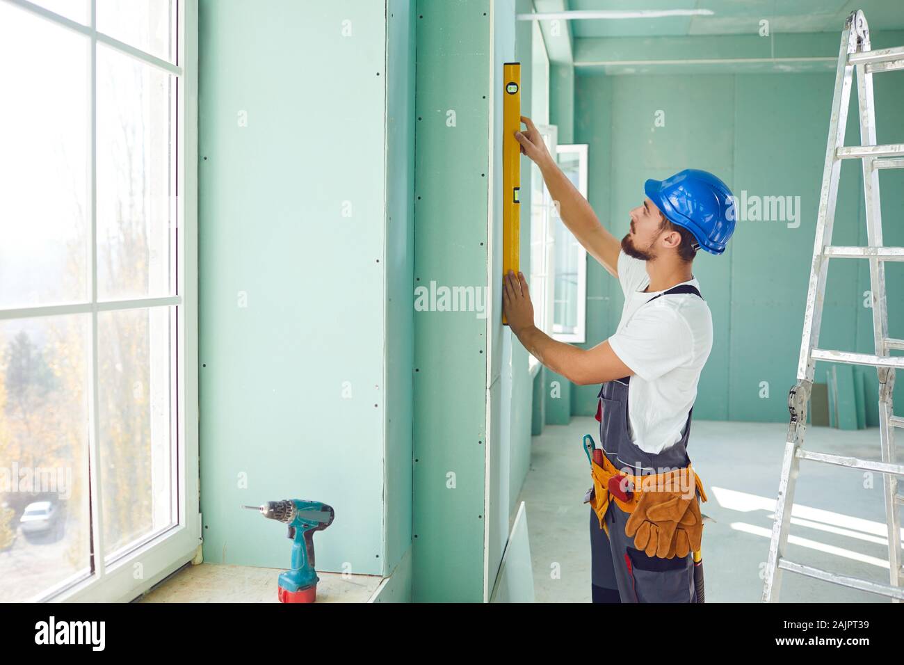 A builder standing on a ladder installs drywall at a construction site ...