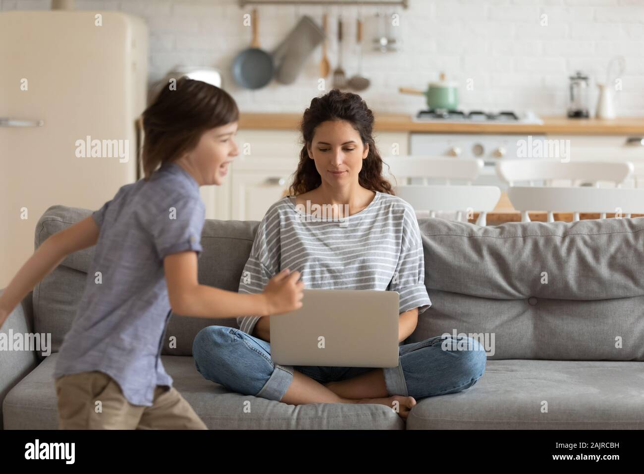 Mother Seated On Couch Using Computer Her Active Son Running Stock