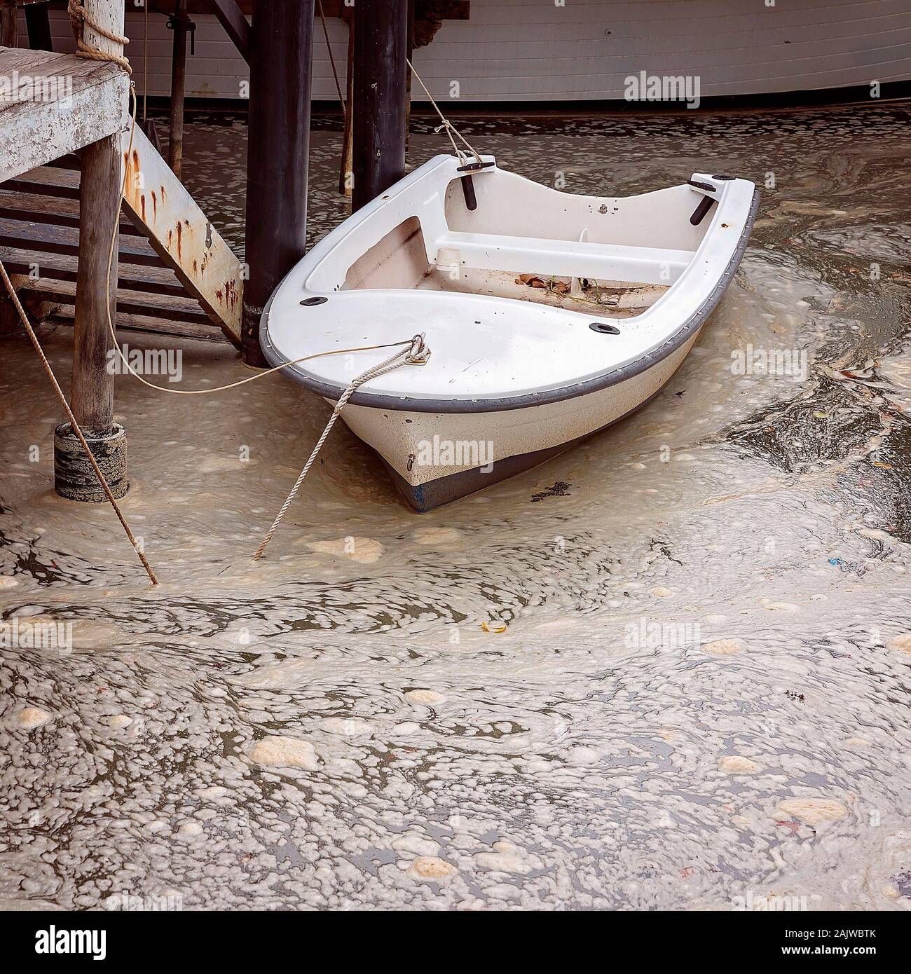 A small white row boat tied up to a post in swirling foamy dirty water ...