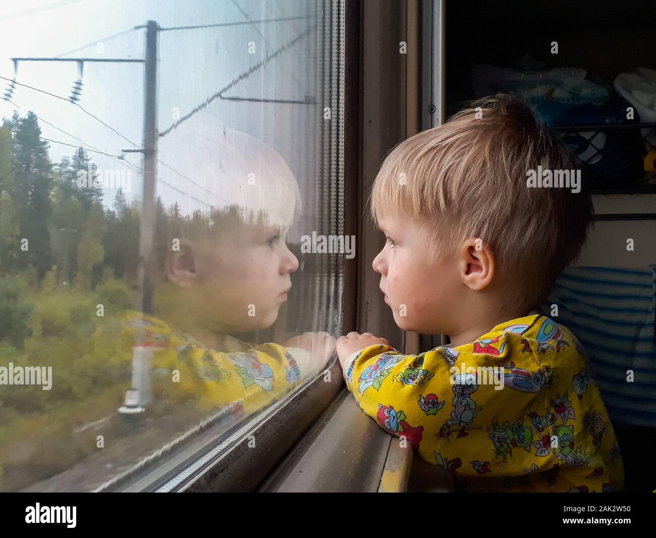 A bright, cute three-year-old boy riding a train looks out the window ...