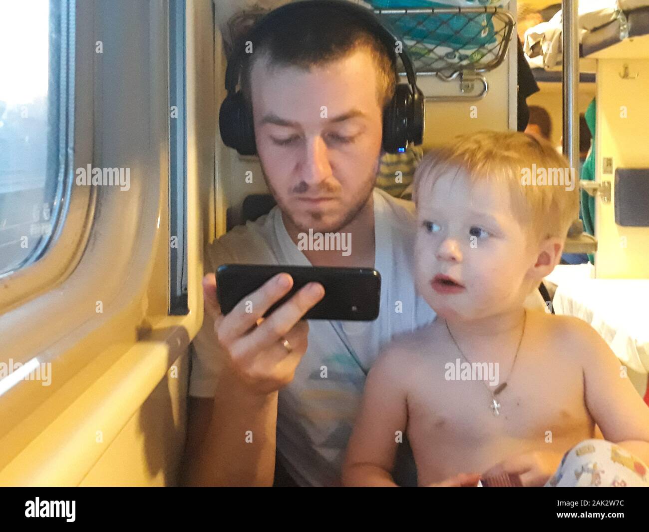 A young father and a young son ride a train. A man with headphones ...