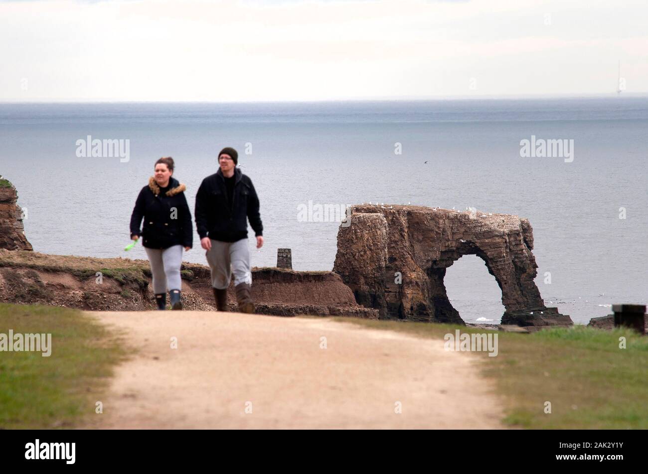 Couple walking with rock arch in the background, Whitburn Coastal Park ...