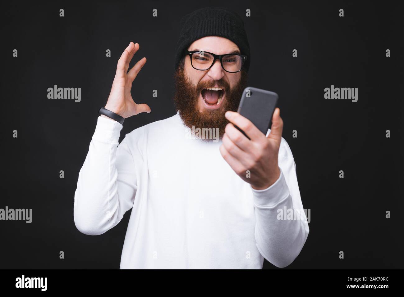 Angry bearded man screaming at smartphone standing over dark background ...