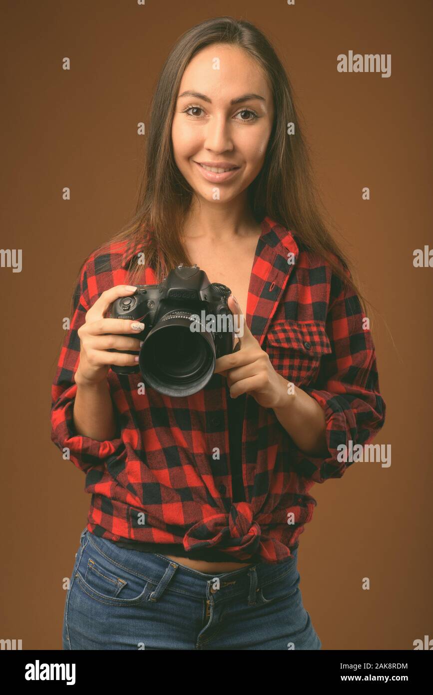 Studio shot of young beautiful woman with camera against brown ...