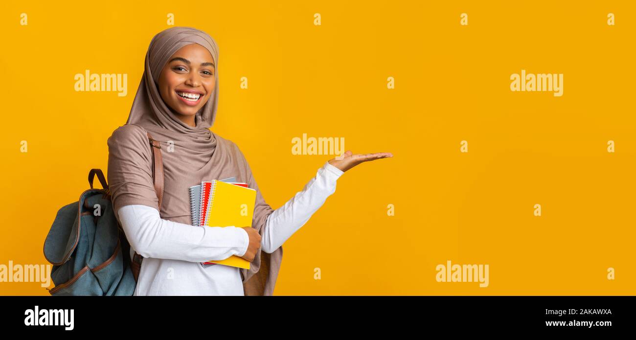 Cheerful black islamic female student in hijab pointing aside at copy ...